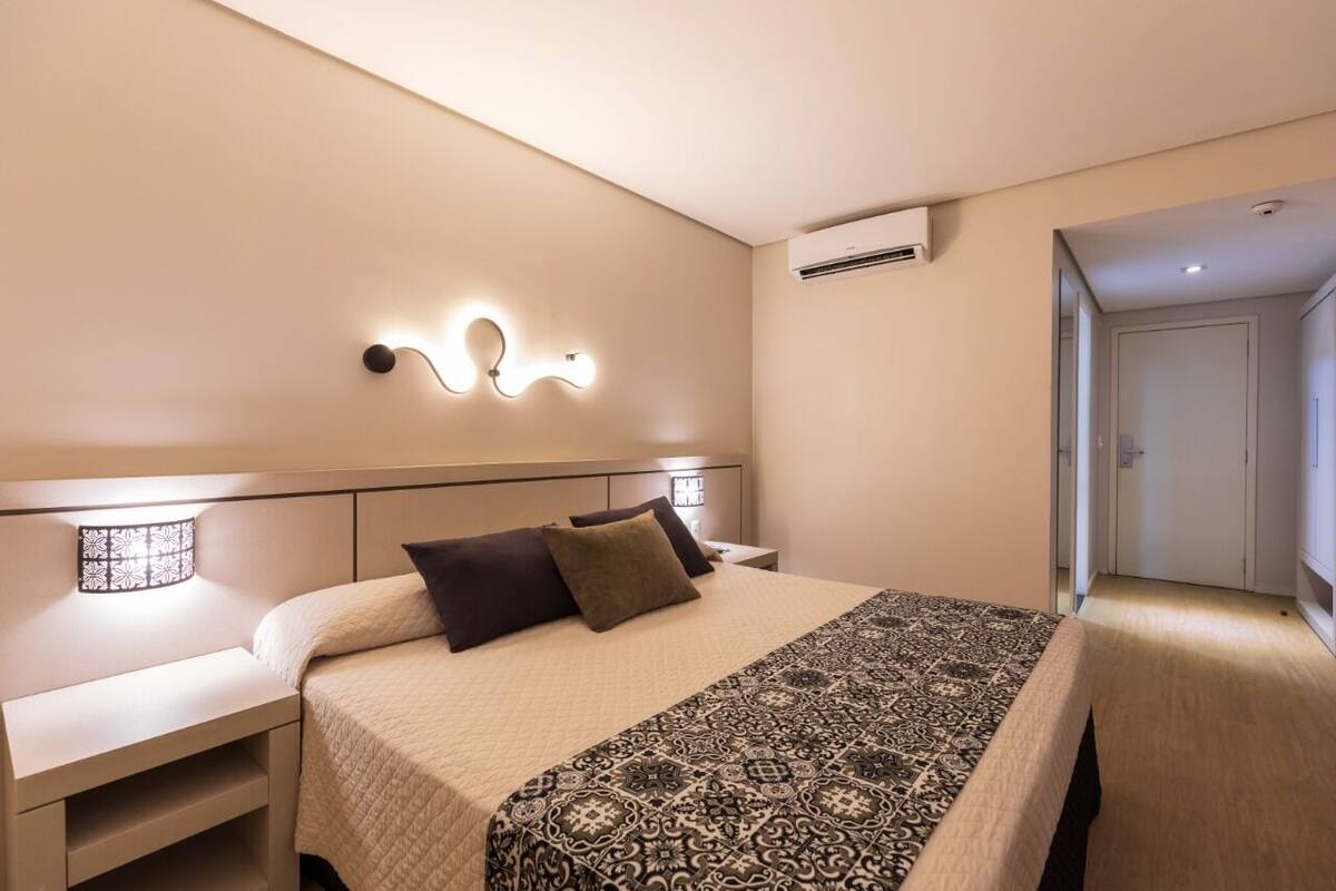 Quarto de hotel moderno com uma cama grande, mesas de cabeceira, luminária fixada na parede e aparelho de ar condicionado.