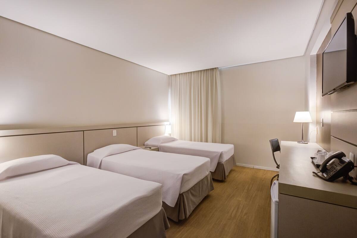 Quarto de hotel com três camas de solteiro, uma escrivaninha com telefone e uma televisão na parede.