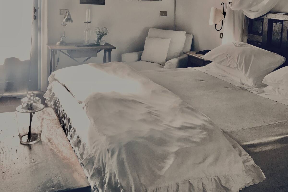 Um quarto com cama, poltrona, mesa de cabeceira, abajur e janela com cortinas.