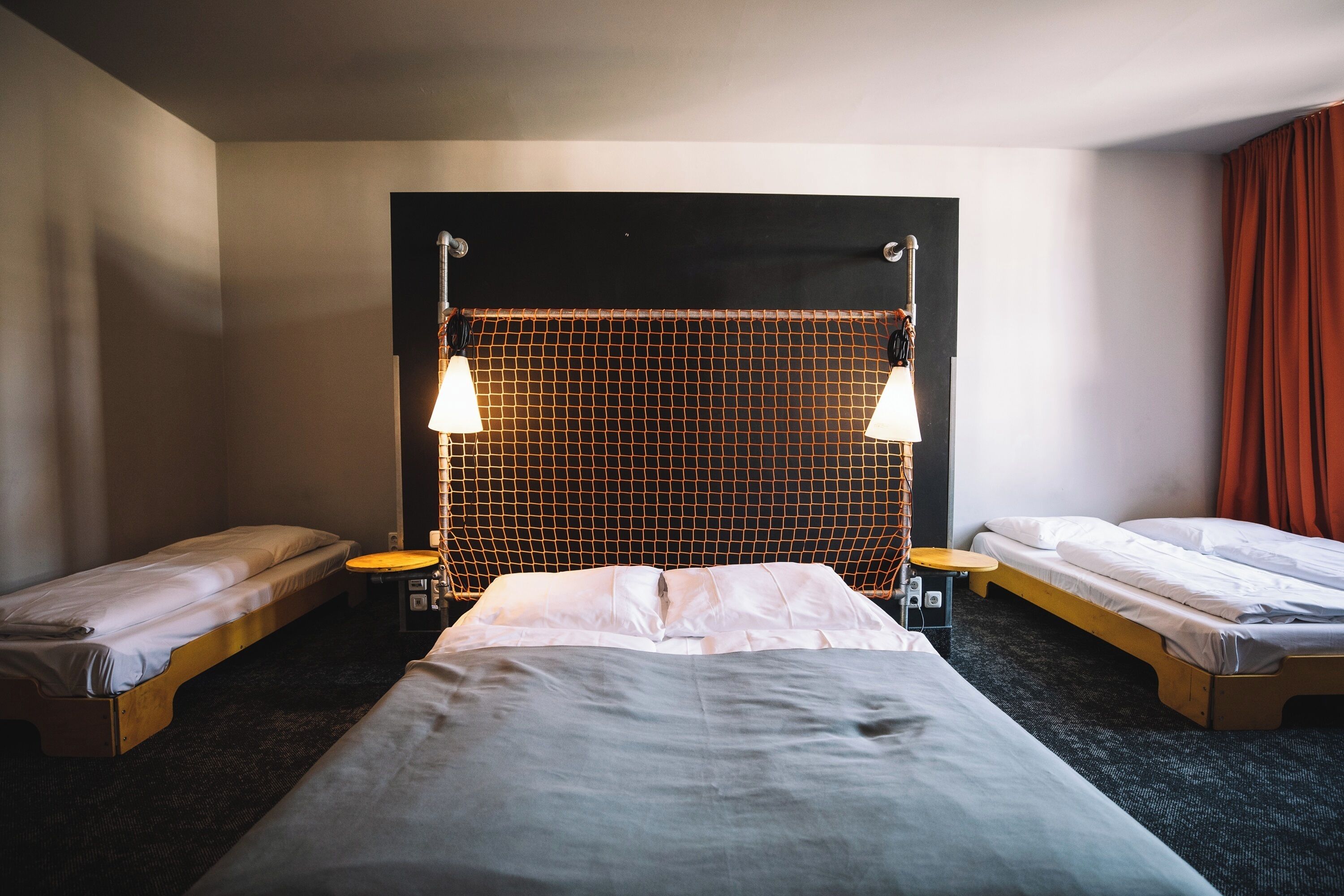 Quarto de hotel com uma cama grande, duas camas menores, uma mesinha e cabeceira com padrão quadriculado.