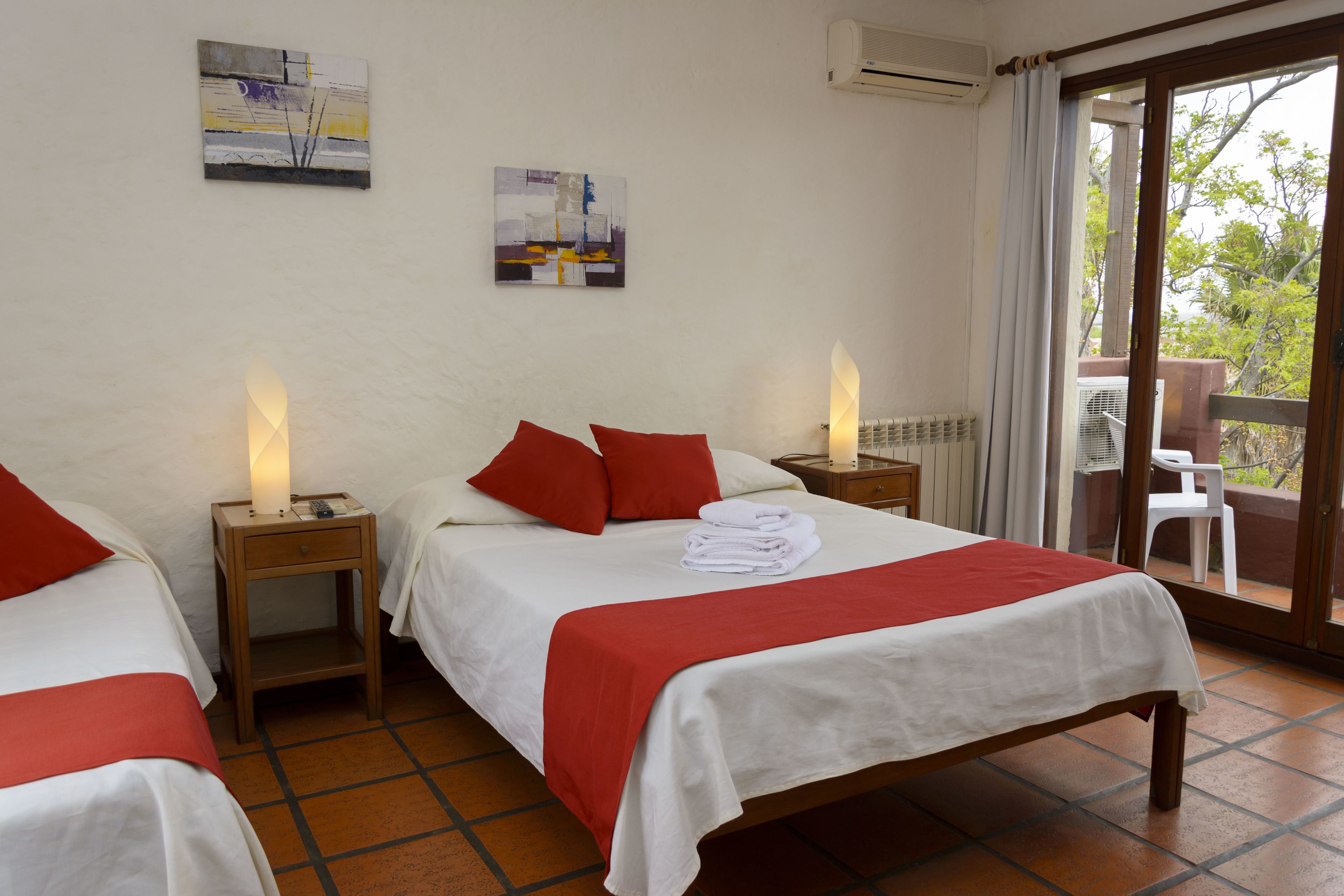 Quarto de hotel com duas camas, cada uma com travesseiro vermelho, roupa de cama branca e uma mesa de cabeceira com abajur. Há duas pinturas penduradas na parede e uma janela com vista para o verde.