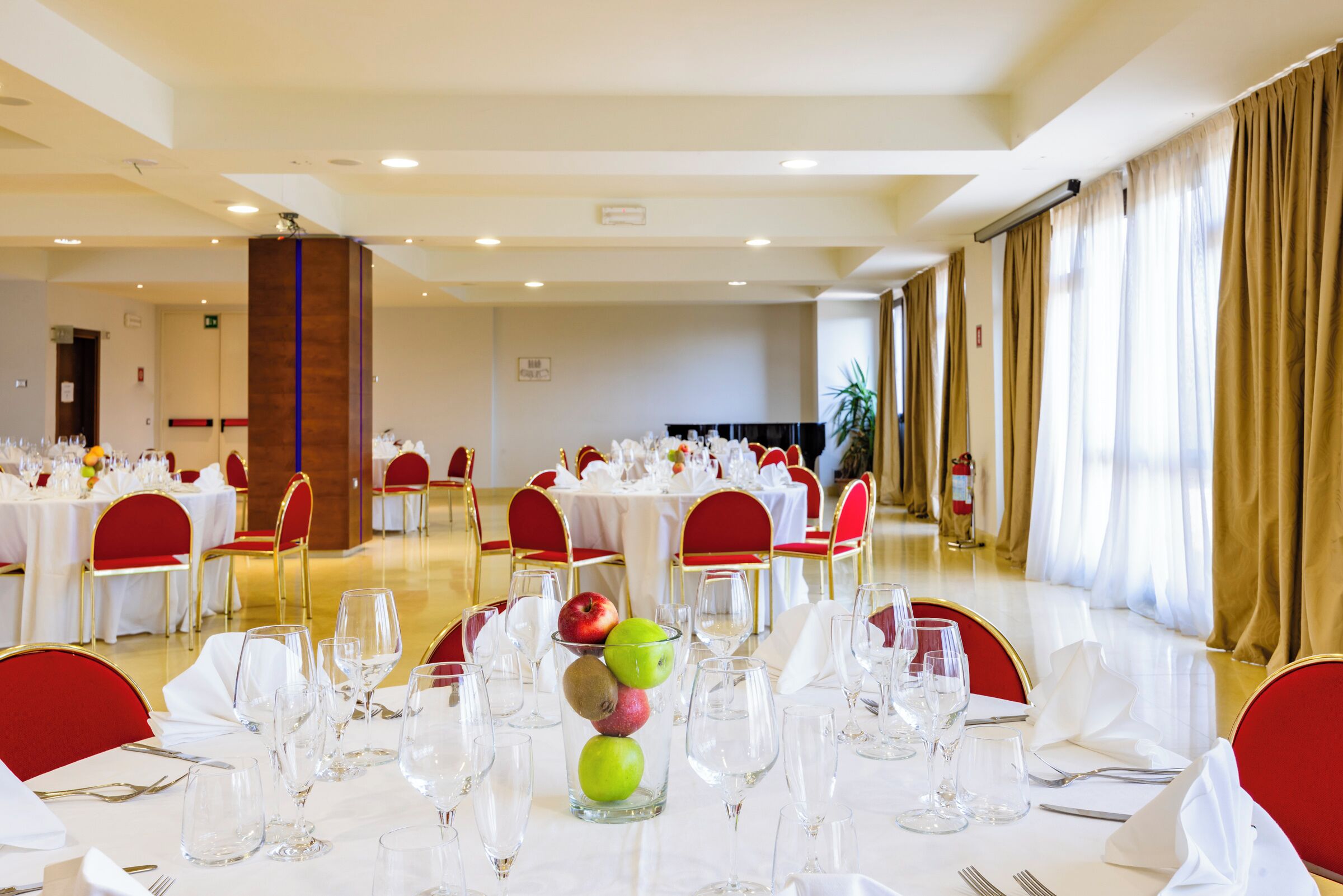 Salão de banquetes com mesas redondas preparadas para refeições, cadeiras vermelhas e um centro de mesa decorativo com frutas.