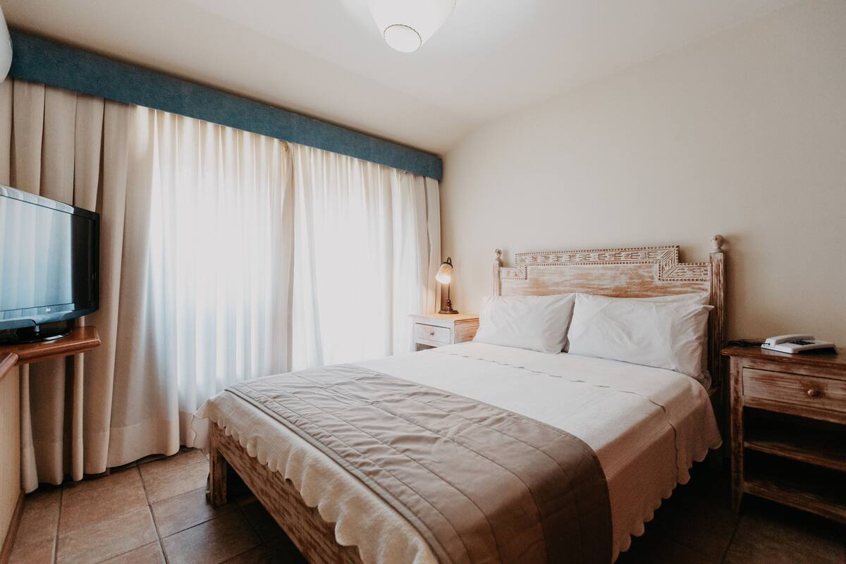 Quarto de hotel com uma cama, televisão sobre um suporte, mesa de cabeceira com abajur e janela com cortinas.