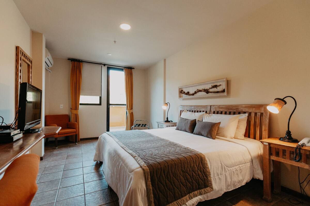 Quarto de hotel com uma cama grande, uma escrivaninha com um computador, uma cadeira e uma televisão.