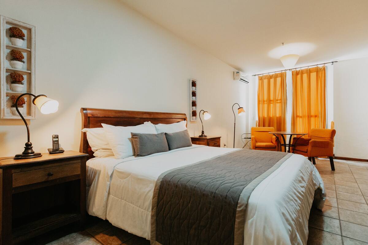 Quarto de hotel com cama, mesas de cabeceira, luminária, cadeira e uma pequena mesa.
