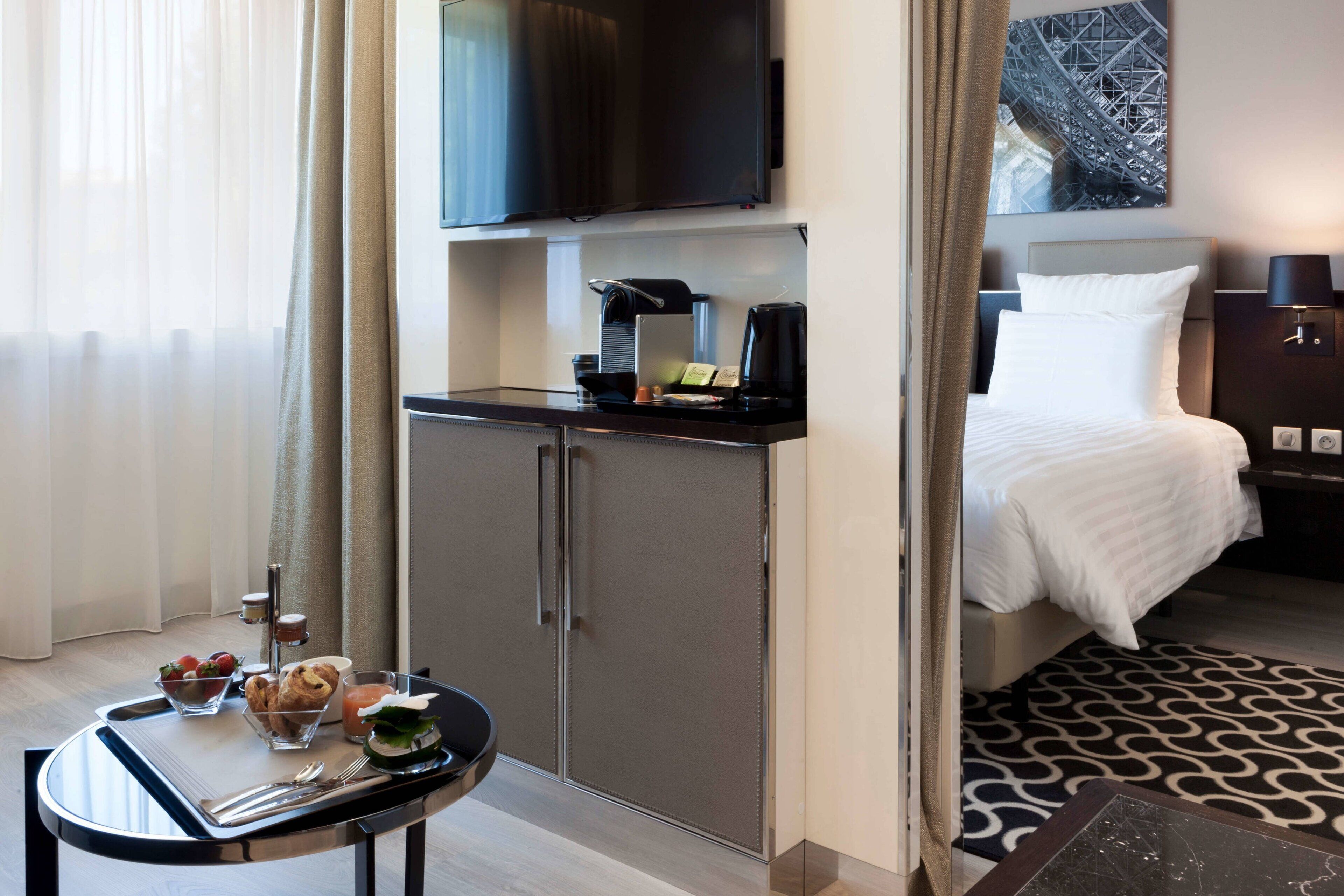 Quarto de hotel com cama, escrivaninha com cafeteira, televisão e área de refeições com uma bandeja de comida.