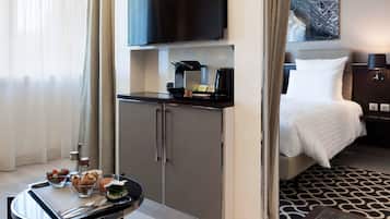 Quarto de hotel com cama, escrivaninha com cafeteira, televisão e área de refeições com uma bandeja de comida.