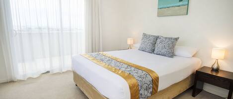 Uma cama bem arrumada com colcha dourada e branca, duas luminárias de cabeceira, um criado-mudo e um quadro com uma paisagem de praia na parede.