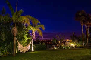 Vista noturna de um jardim tropical com rede, palmeiras e uma paisagem urbana ao longe.