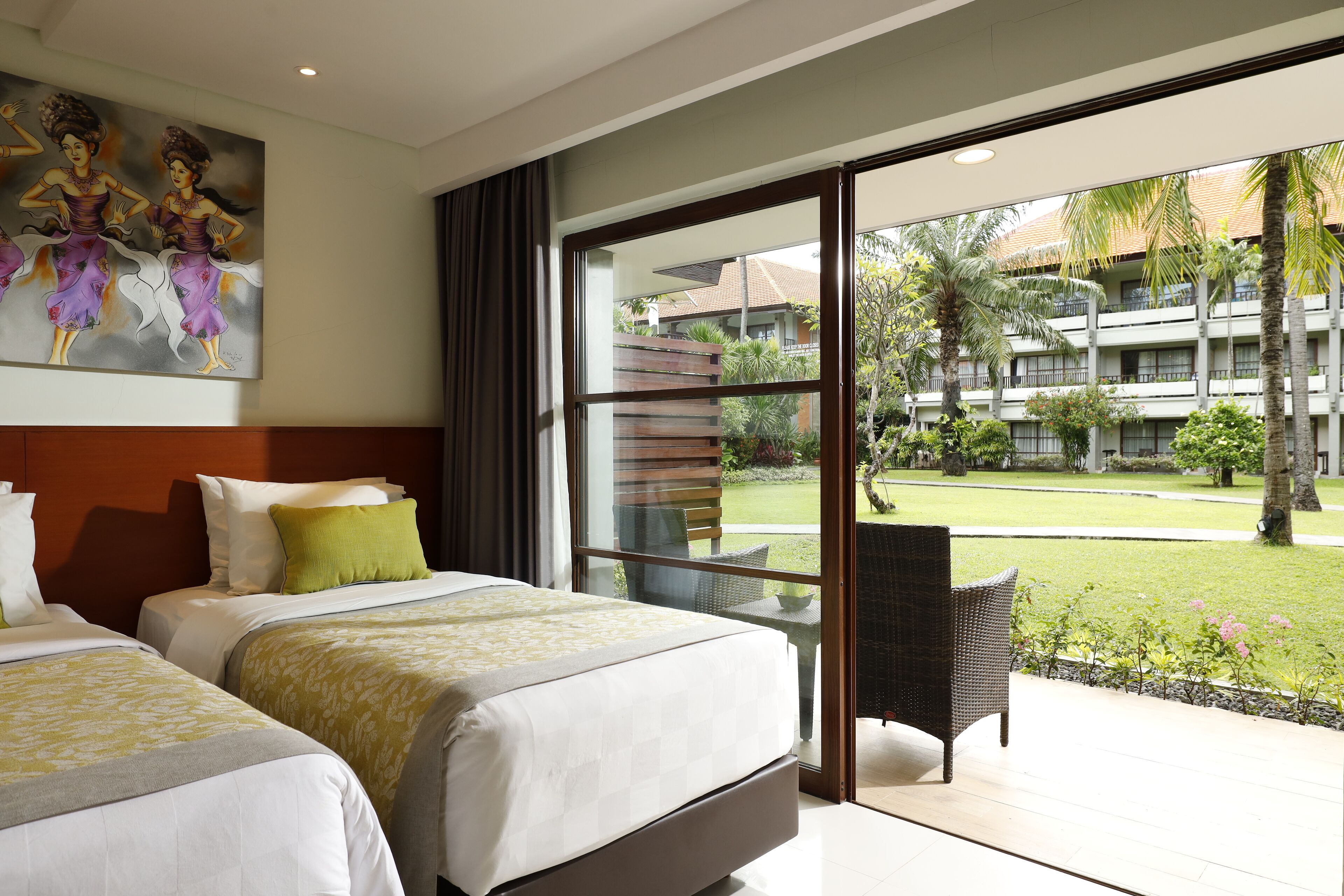 Quarto de hotel com duas camas, uma janela grande e vista para um pátio com palmeiras.