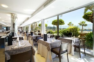 Restaurante com mesas preparadas para refeições, amplas janelas com vista para a praia e vegetação.