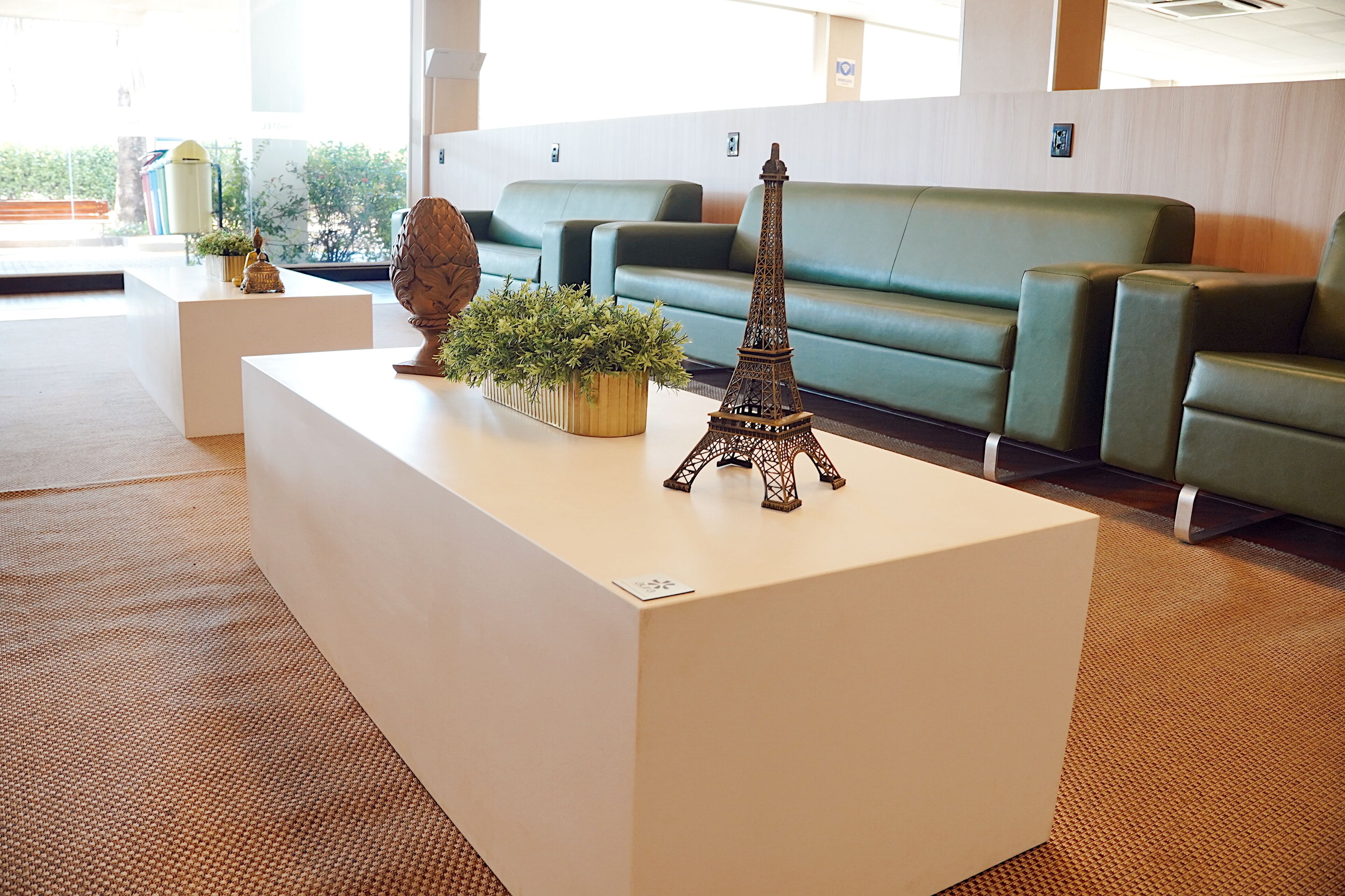 Uma área de recepção com um balcão branco, um sofá verde, uma pequena planta em vaso e uma réplica decorativa da Torre Eiffel.
