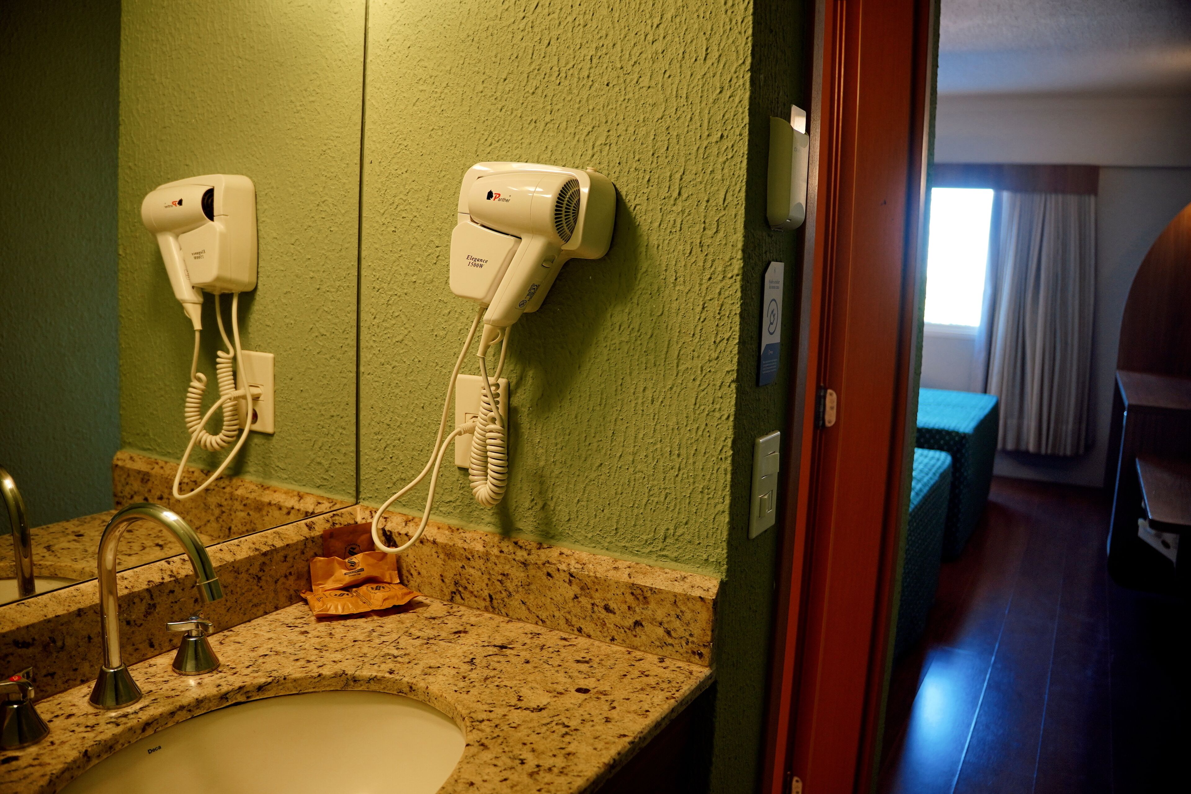 Casa de banho com bancada de granito, lavatório e dois secadores de cabelo fixados na parede.
