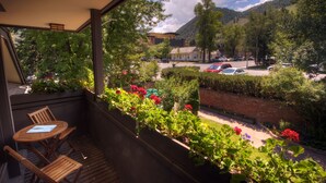 Uma varanda com cadeiras e mesa de madeira, com vista para uma rua com carros e edifícios, e uma montanha ao fundo.