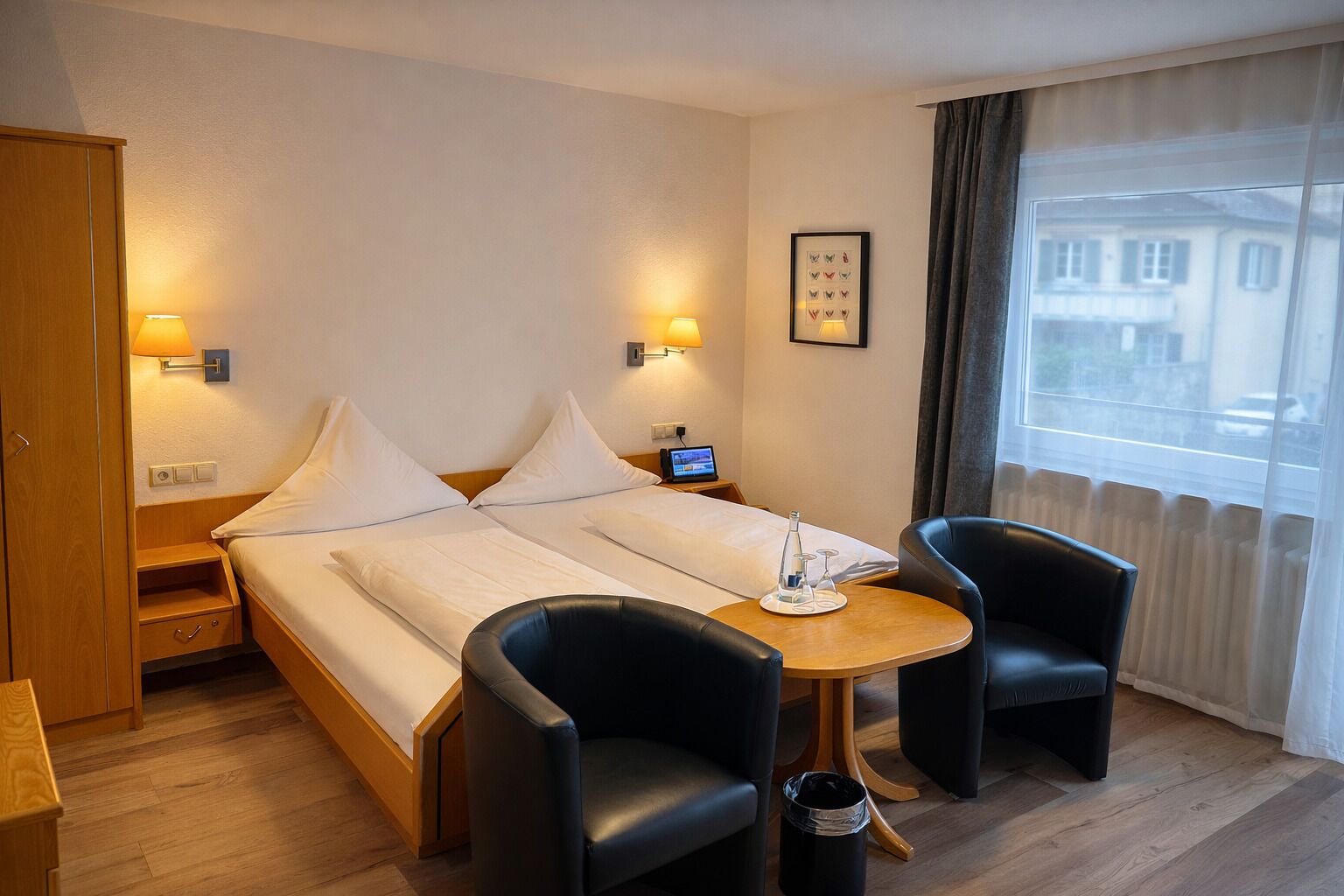 Quarto de hotel com duas camas, uma mesa pequena com cadeiras, uma mesa de cabeceira, um abajur e uma janela com cortinas.