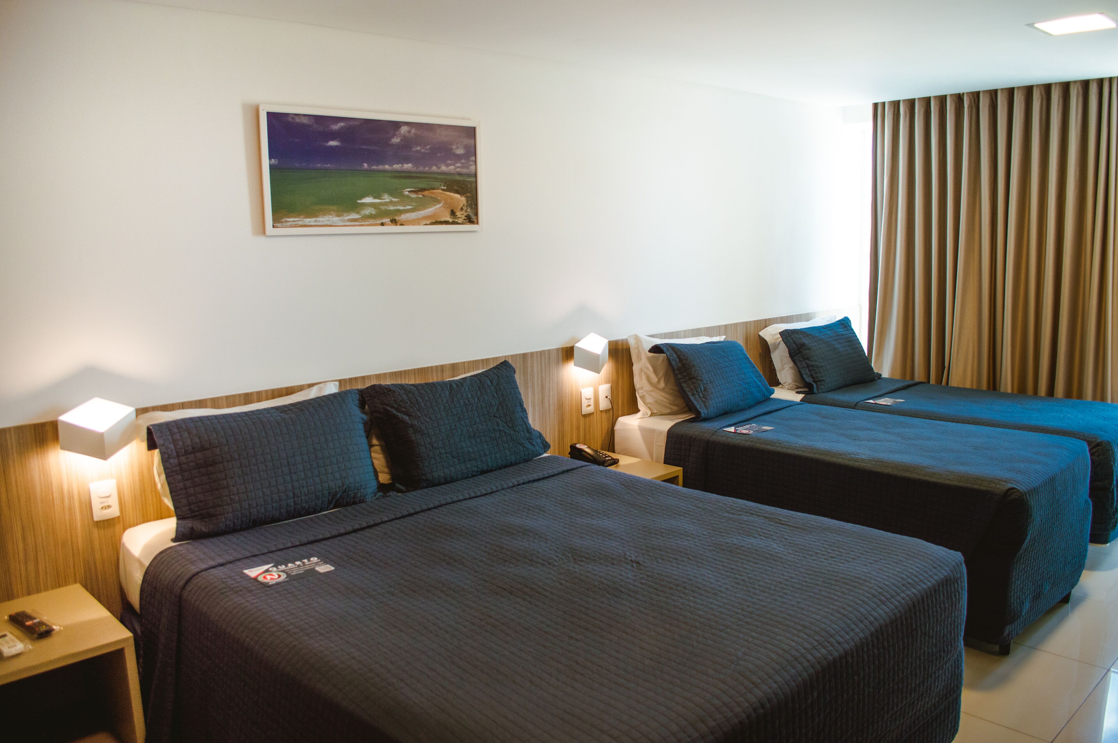 Quarto de hotel com duas camas, uma mesa de cabeceira, um quadro de uma praia e uma cortina.