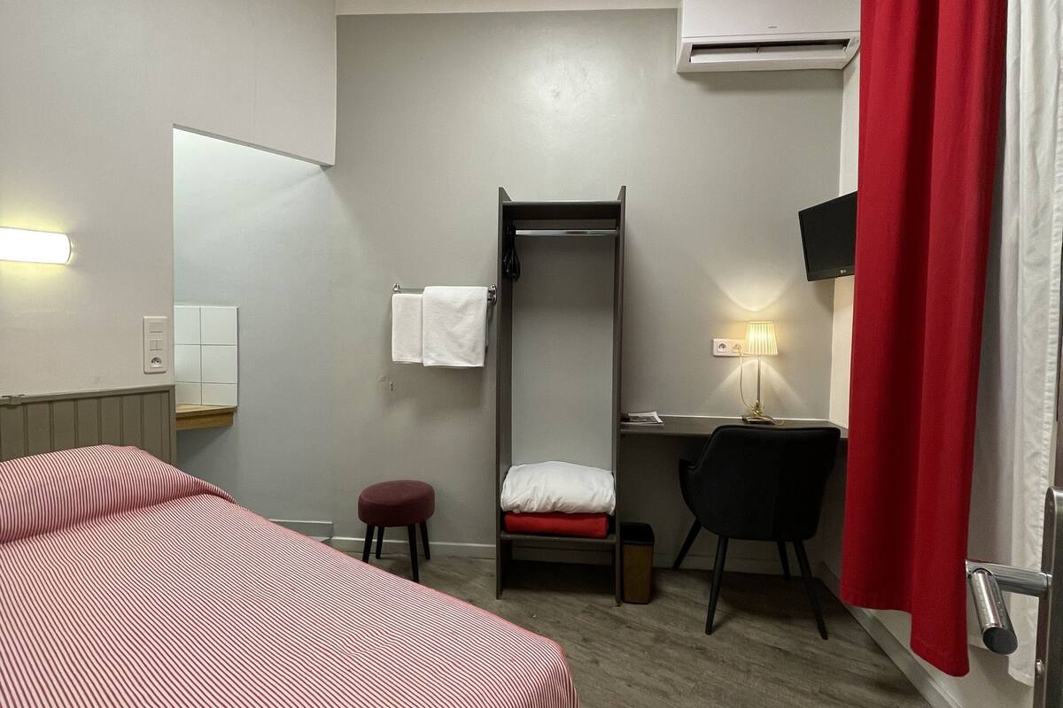 Quarto de hotel com cama, escrivaninha, cadeira, espelho e televisão.