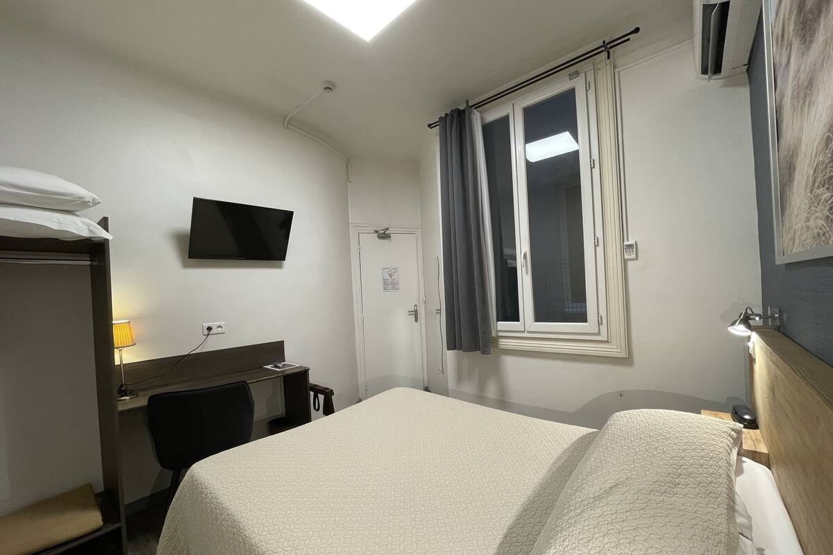 Quarto de hotel com cama, escrivaninha, cadeira, televisão, janela e cortinas.