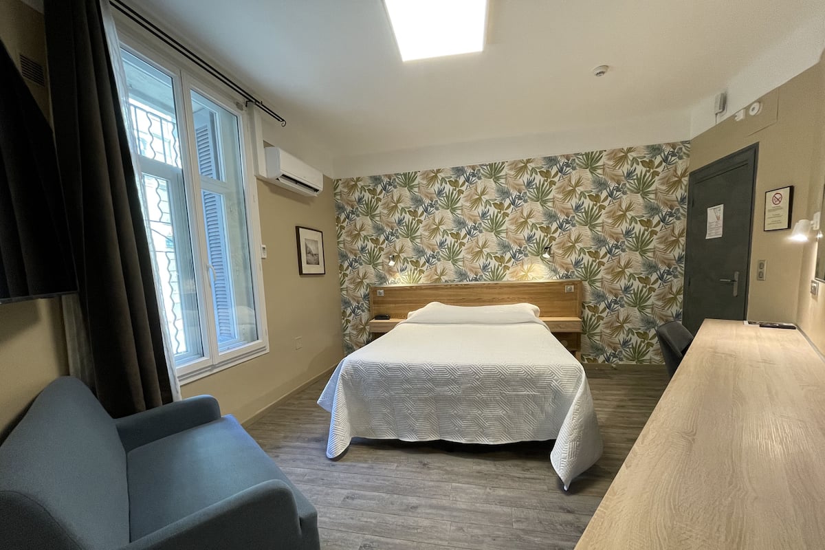 Quarto de hotel com cama, escrivaninha, cadeira, janela com persianas e parede com padrão de folhagem tropical.