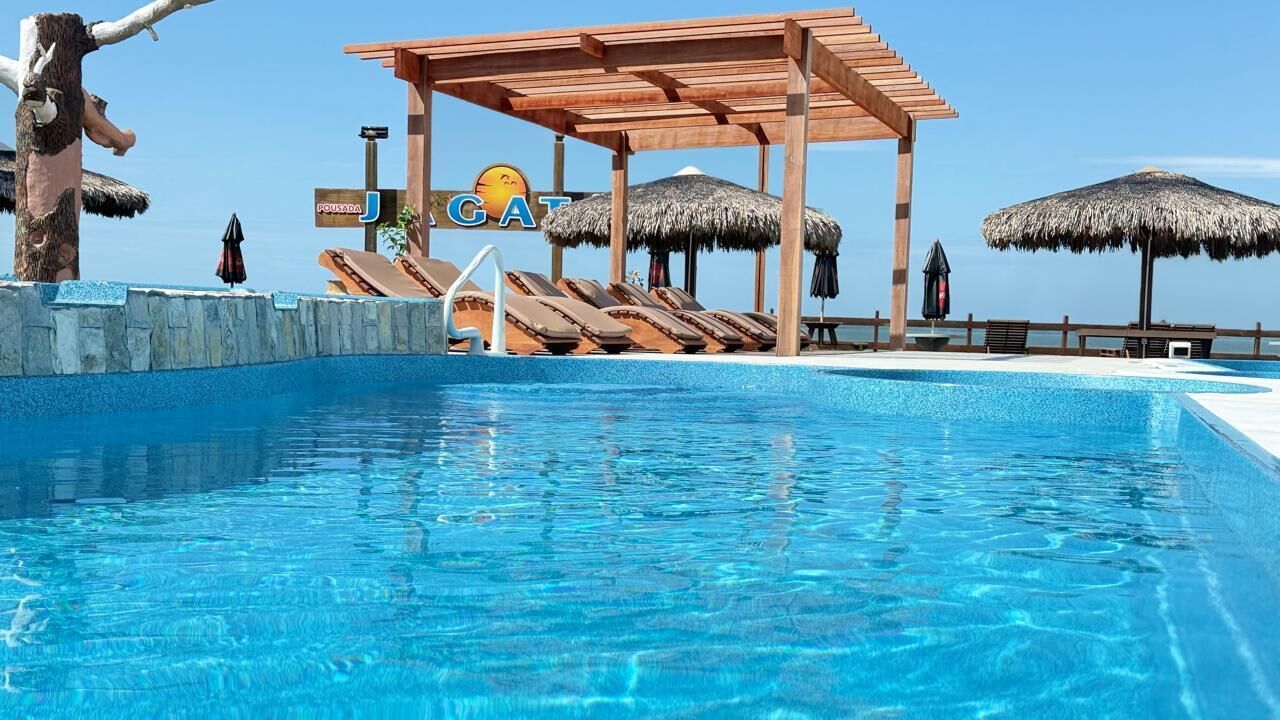 Piscina azul límpida com espreguiçadeiras de madeira e guarda-sóis de palha, uma pérgula de madeira e uma placa com 'Foukas' e 'GAT' visíveis.