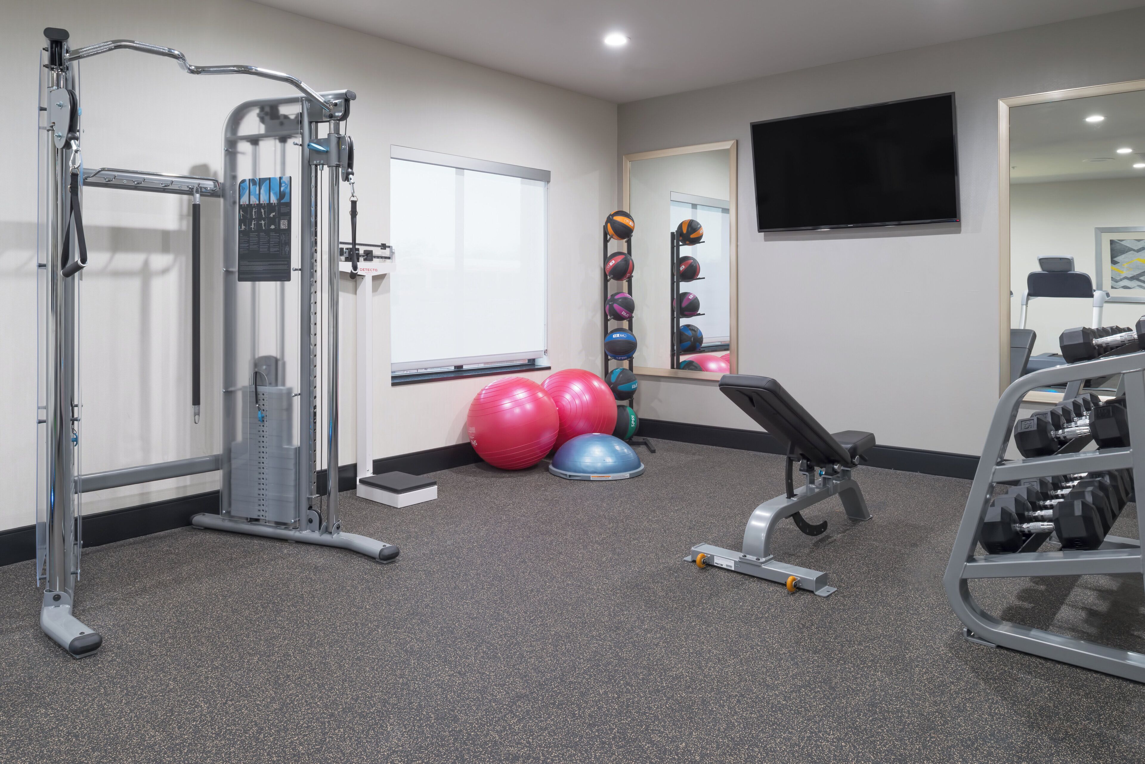 Academia com equipamentos de ginástica, televisão presa à parede e espelho.