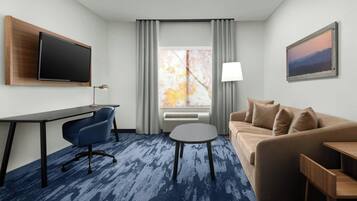 Quarto de hotel com televisão de tela plana, escrivaninha, cadeira, sofá, mesa de centro, abajur e um quadro na parede.