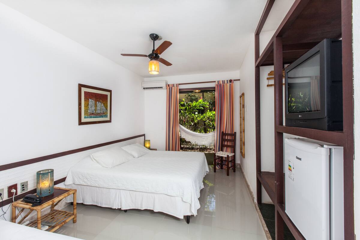 Quarto de hotel com uma cama, televisão, ventoinha no teto, uma mesinha com luminária e uma janela com cortinas.
