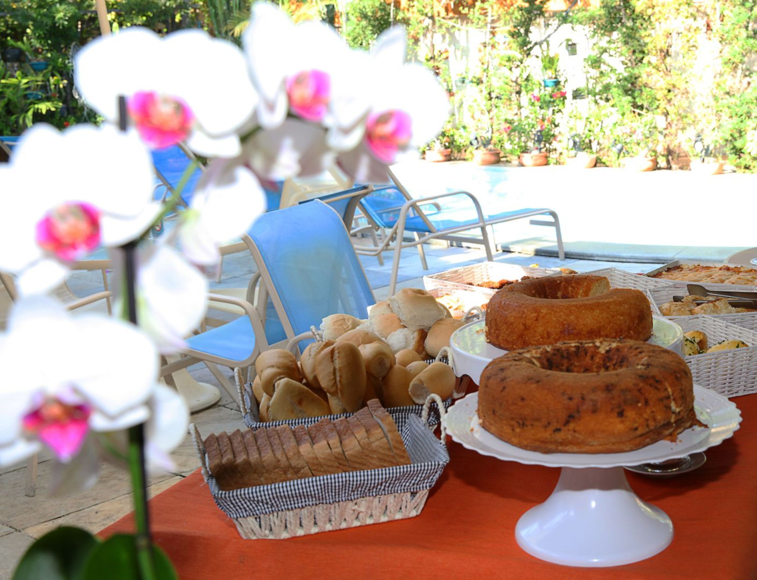 Uma mesa com diversos produtos de pastelaria e um bolo em suporte, rodeada por cadeiras e uma piscina ao fundo.