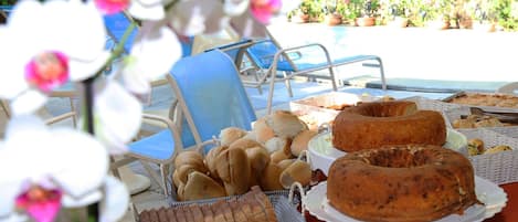 Uma mesa com diversos produtos de pastelaria e um bolo em suporte, rodeada por cadeiras e uma piscina ao fundo.
