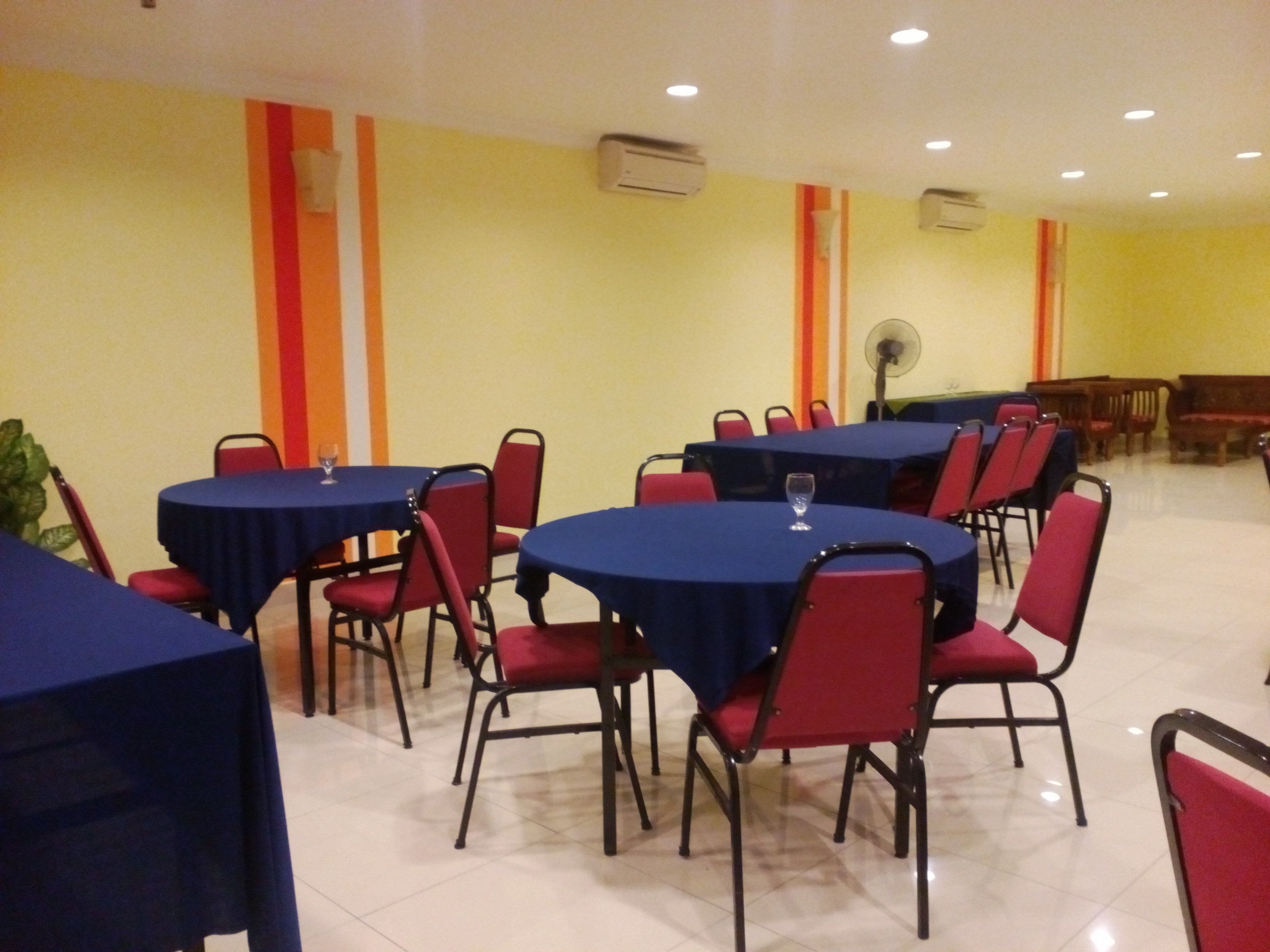 Uma área de refeições com mesas redondas cobertas por toalhas azuis, cadeiras vermelhas e um ventilador na parede.