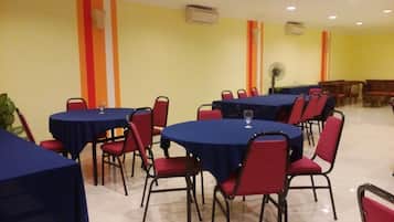 Uma área de refeições com mesas redondas cobertas por toalhas azuis, cadeiras vermelhas e um ventilador na parede.