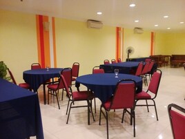 Uma área de refeições com mesas redondas cobertas por toalhas azuis, cadeiras vermelhas e um ventilador na parede.