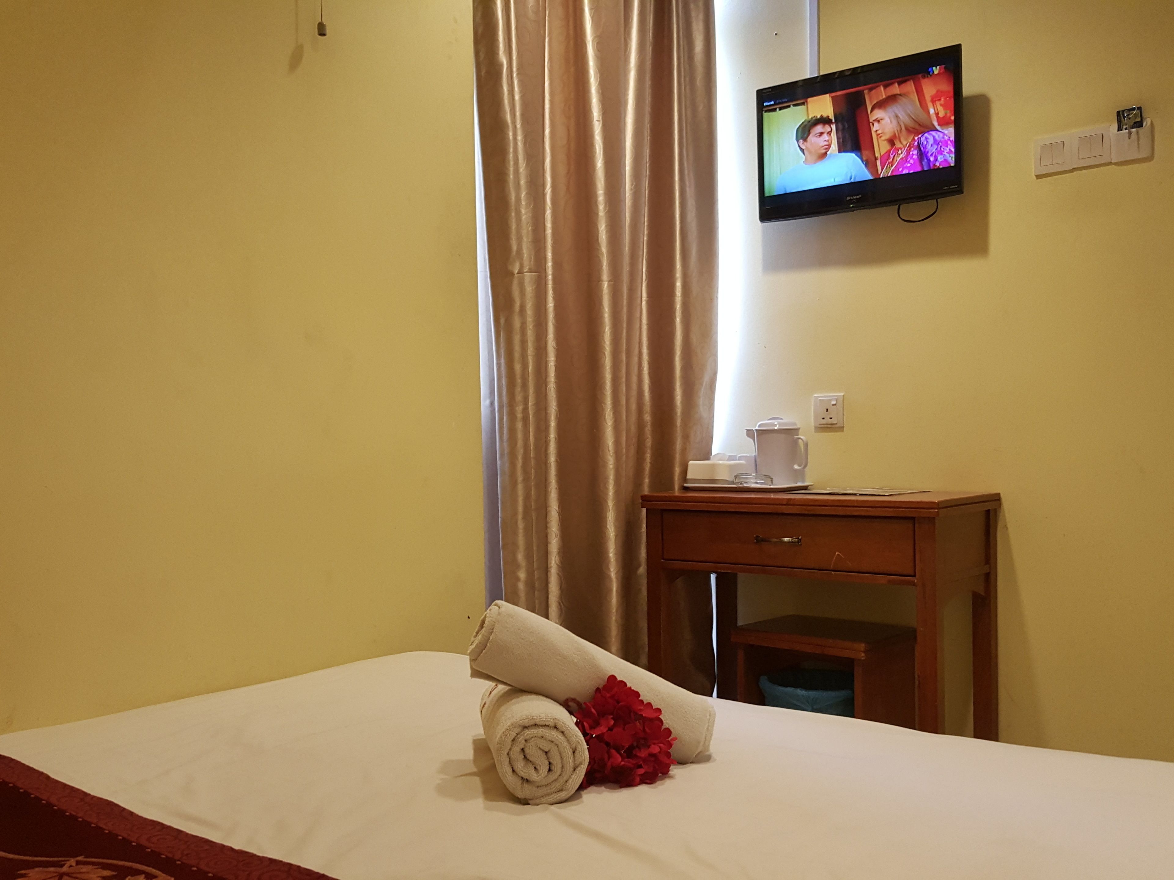 Quarto de hotel com cama, televisão fixada na parede, mesa de cabeceira de madeira e janela com cortinas.