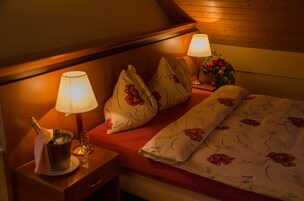 Quarto de hotel com cama, duas luminárias de cabeceira, mesa de cabeceira com uma garrafa de champanhe e uma taça, e colcha com padrão floral.
