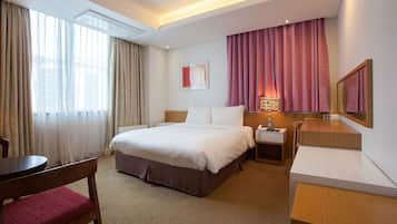 Quarto de hotel com uma cama grande, uma escrivaninha, uma cadeira, mesa de cabeceira, abajur e espelho.