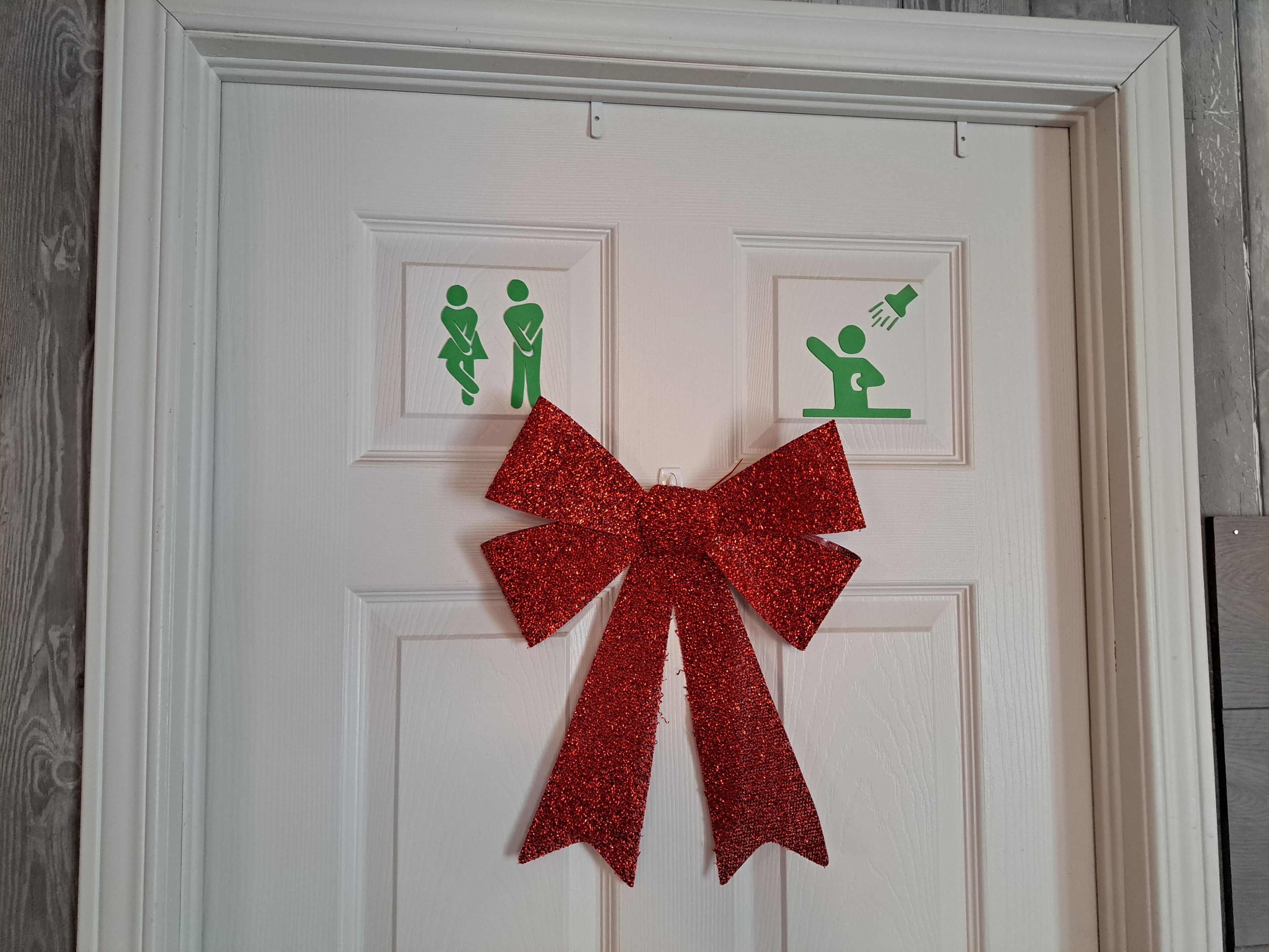 Uma porta branca com um laço vermelho brilhante e duas placas verdes indicando banheiros.