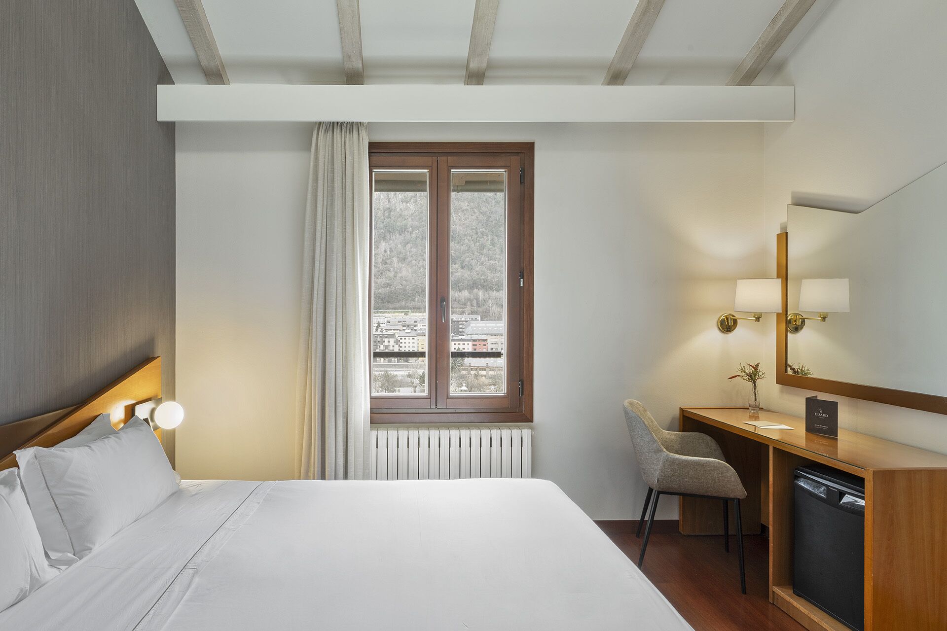 Quarto de hotel com cama, secretária com cadeira, janela com cortinas, radiador e espelho com iluminação.