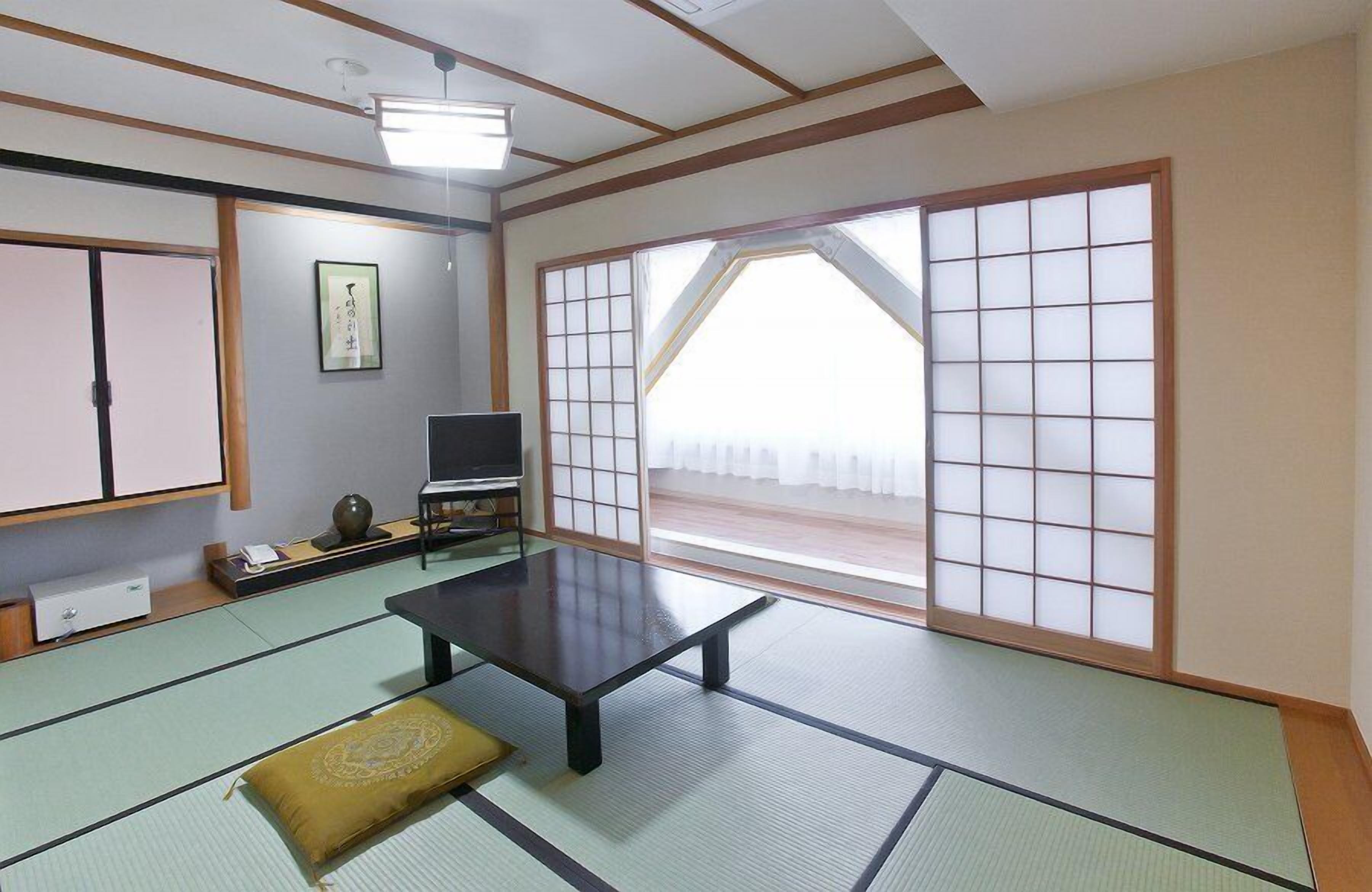 Um quarto japonês tradicional com tatames, uma mesa baixa, portas deslizantes, uma televisão e uma obra de caligrafia emoldurada.
