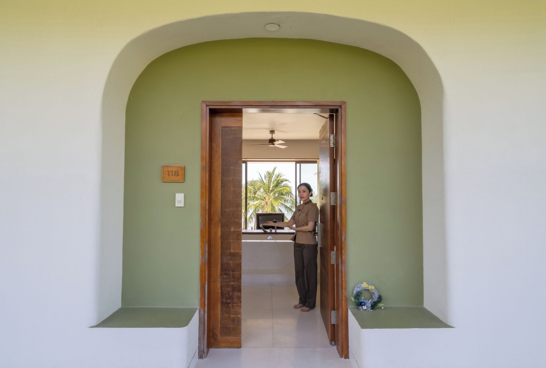Área de recepção de hotel com recepcionista, balcão e vista para palmeiras.