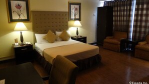 Quarto de hotel com uma cama grande, duas mesas de cabeceira com luminárias, uma escrivaninha e uma cadeira. Há duas pinturas florais emolduradas nas paredes.