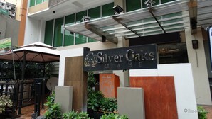 Exterior do Hotel Silver Oaks com design moderno, apresentando uma placa de destaque, grandes janelas e uma área de estar externa coberta.