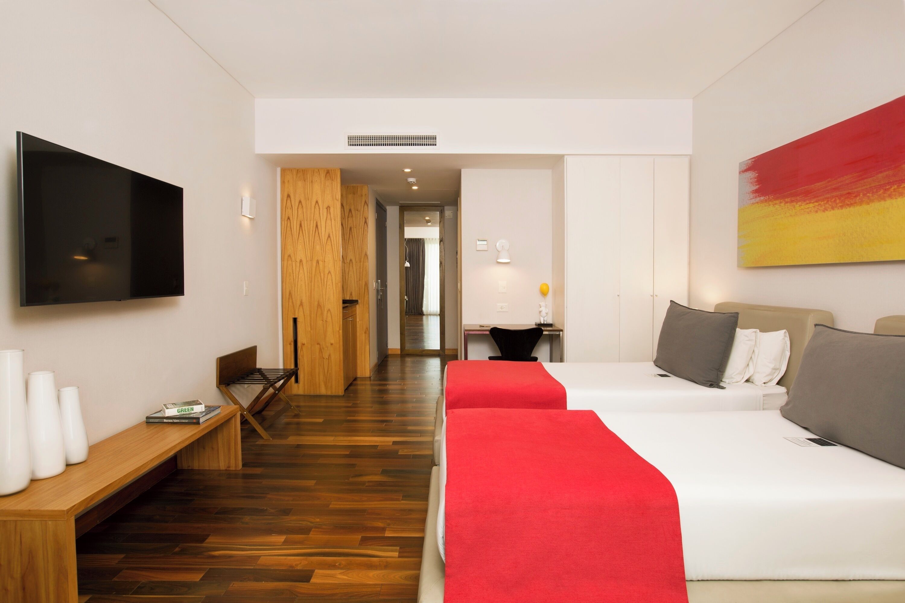 Quarto de hotel com uma grande televisão de tela plana, um banco de madeira, uma escrivaninha com cadeira e uma cama com roupa de cama vermelha e cinza.