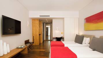 Quarto de hotel com uma grande televisão de tela plana, um banco de madeira, uma escrivaninha com cadeira e uma cama com roupa de cama vermelha e cinza.