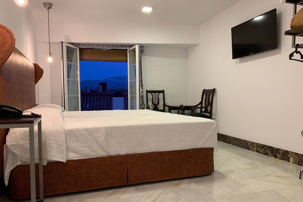 Quarto de hotel com cama, escrivaninha com telefone, cadeira, mesa, TV fixada na parede e janela com vista para edifícios.