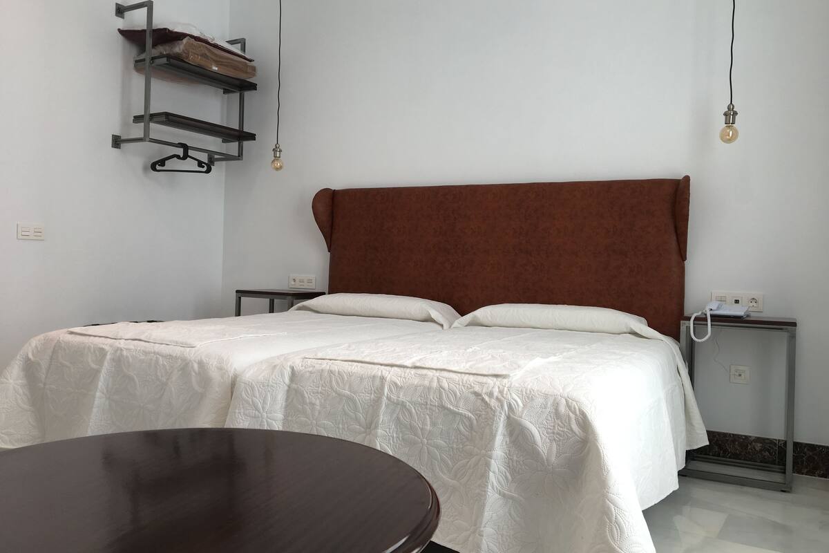 Quarto com cama, mesas de cabeceira, escrivaninha e luminária suspensa.