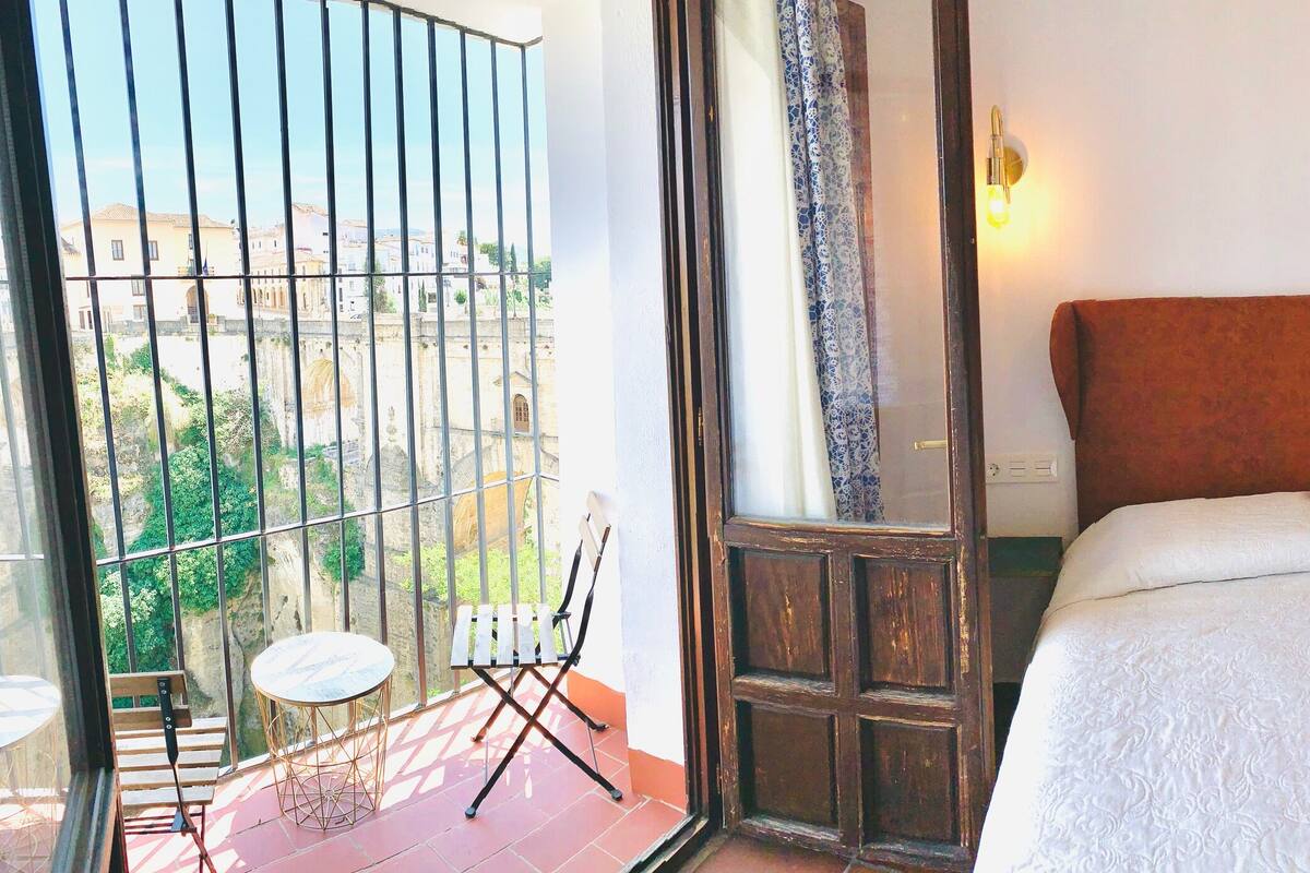 Um quarto com uma cama, uma janela com vista para prédios, uma porta e uma pequena mesa com uma cadeira.