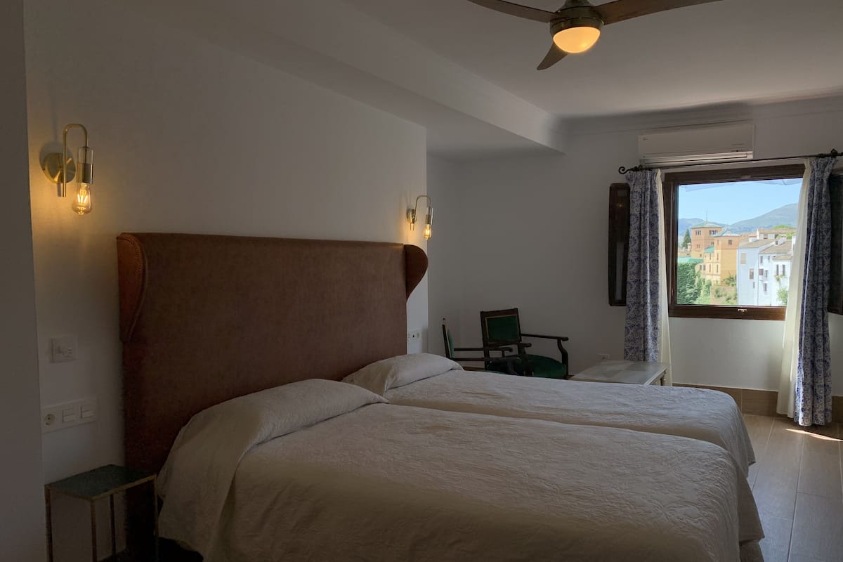 Quarto de hotel com duas camas, ventoinha no teto, janela com cortinas e uma cadeira.
