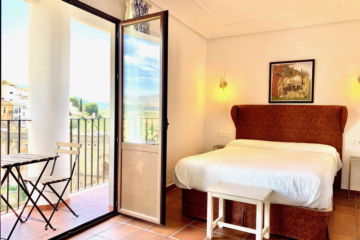 Quarto com cama, uma mesinha, cadeira, varanda e um quadro na parede.