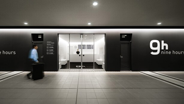 Entrada moderna de hotel com design minimalista, exibindo uma grande placa '9h nine hours' nas paredes, piso de azulejos limpo e uma pessoa caminhando em direção à entrada.