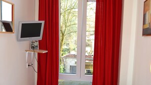 Quarto de hotel com cama, cortinas vermelhas, televisão, pequena mesa com um vaso de flores e janela com vista para árvores.